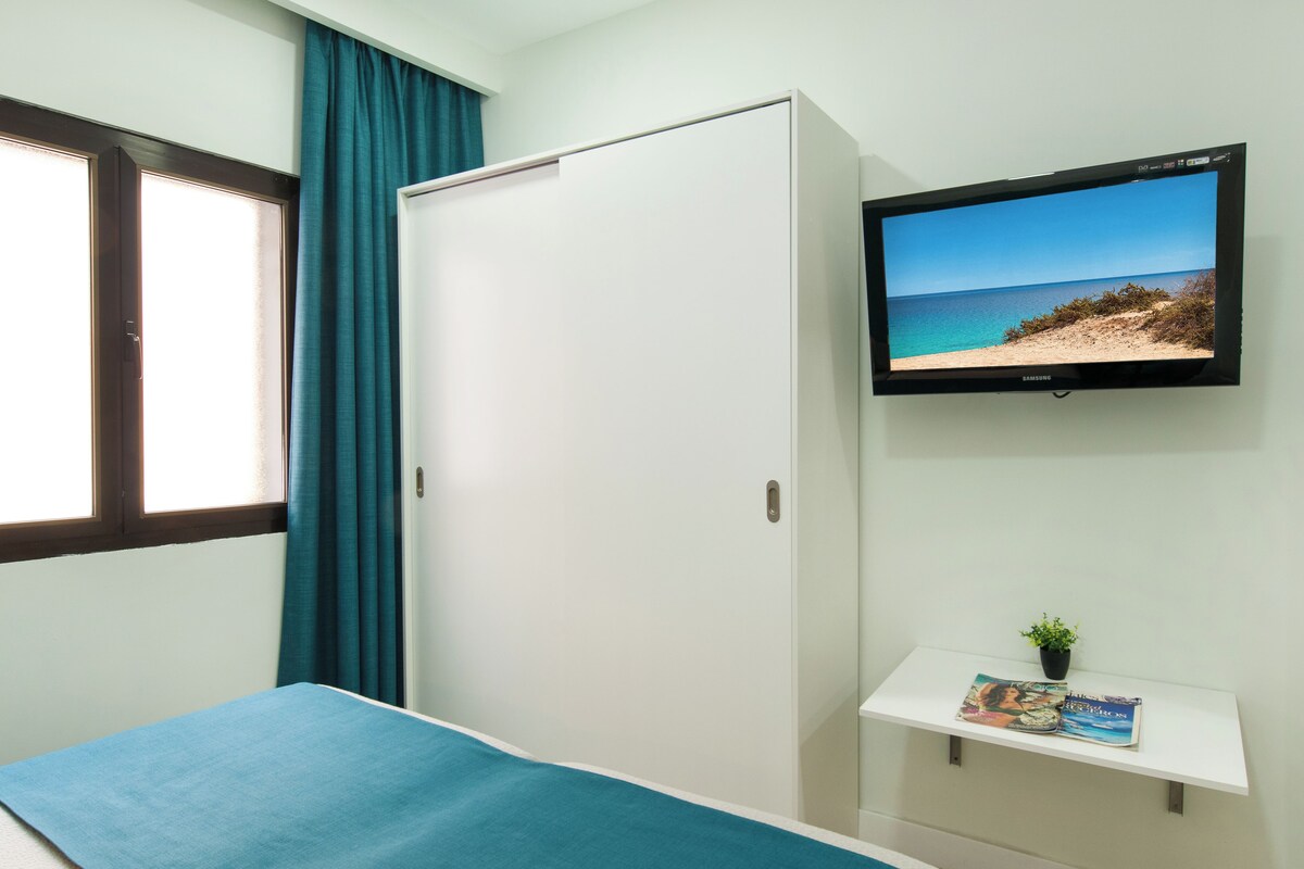 Quarto com cama, guarda-roupa, televisão mostrando uma cena de praia, prateleira pequena com uma planta e janela com cortinas.