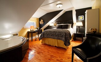 Quarto com uma cama grande, área de estar com uma poltrona, escrivaninha, abajur e banheiro com banheira.