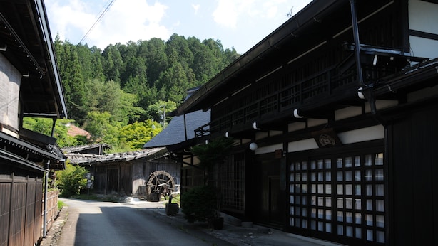 Uma rua tradicional japonesa com edifícios de madeira, uma roda d'água e uma encosta arborizada ao fundo.