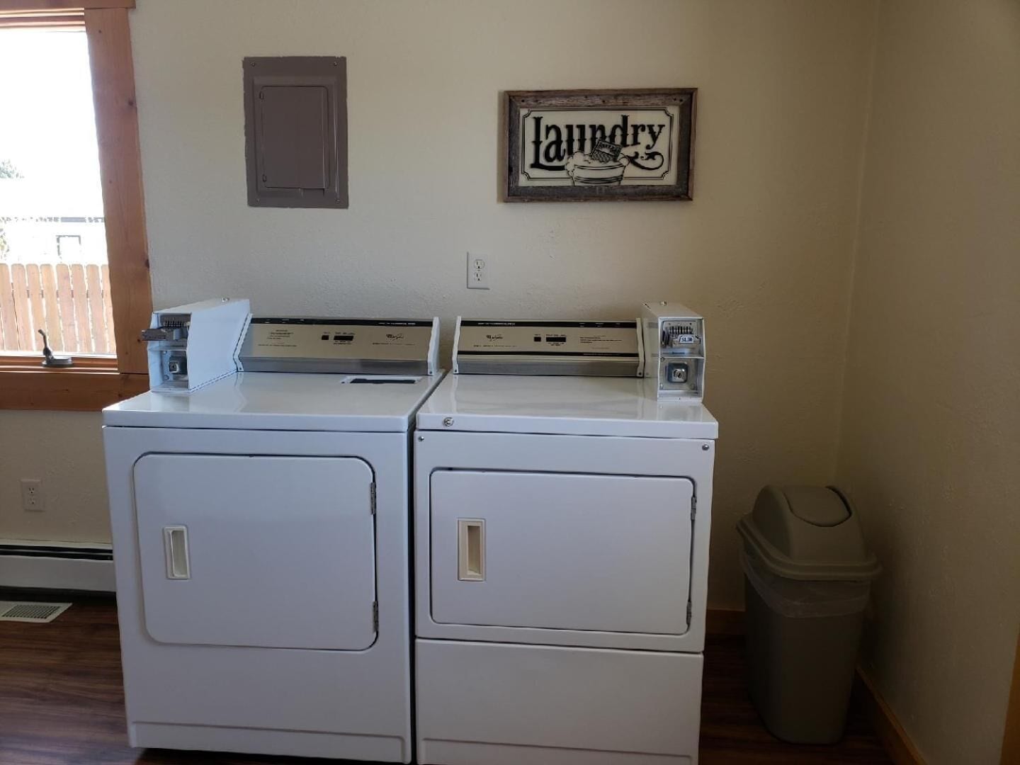 Uma lavanderia com duas máquinas de lavar e secar de carregamento frontal, piso de madeira, janela com persianas e uma placa indicando 'Laundry'.