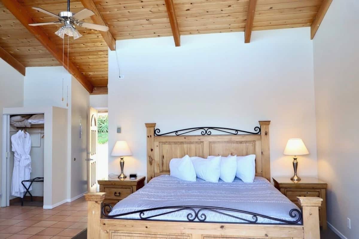 Quarto com estrutura de cama de madeira, duas mesas de cabeceira com luminárias, um ventilador de teto e teto de madeira.