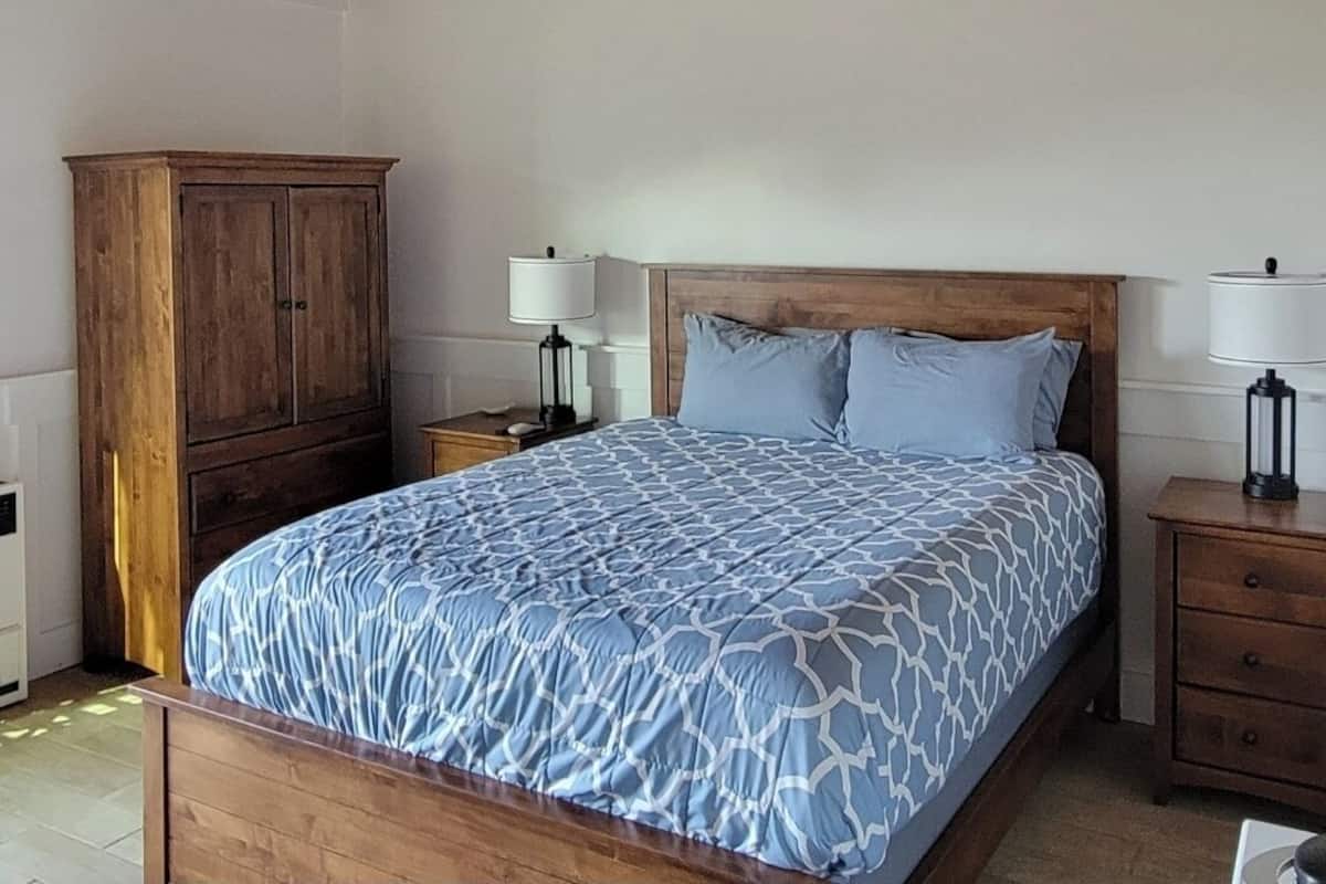 Quarto com teto de madeira, cama com colcha azul e branca estampada, duas mesas de cabeceira com luminárias e um guarda-roupa de madeira.