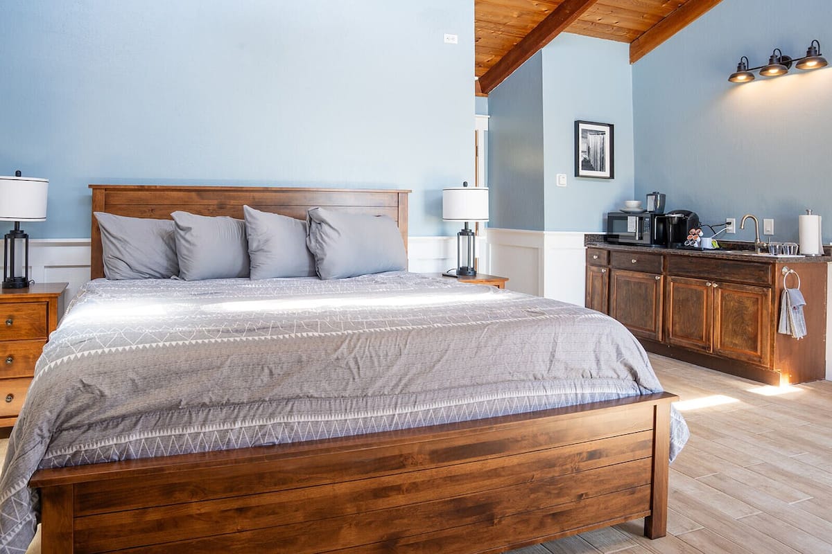Quarto com cama de madeira, duas mesas de cabeceira com luminárias, guarda-roupa de madeira e uma cozinha compacta no canto.