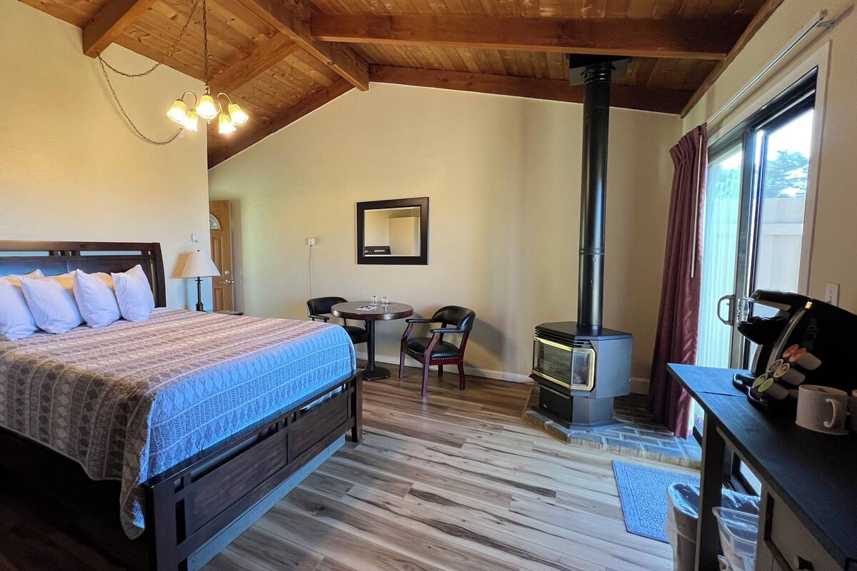 Quarto com cama, teto de madeira, mesa com cadeiras, lareira e janela com cortinas.