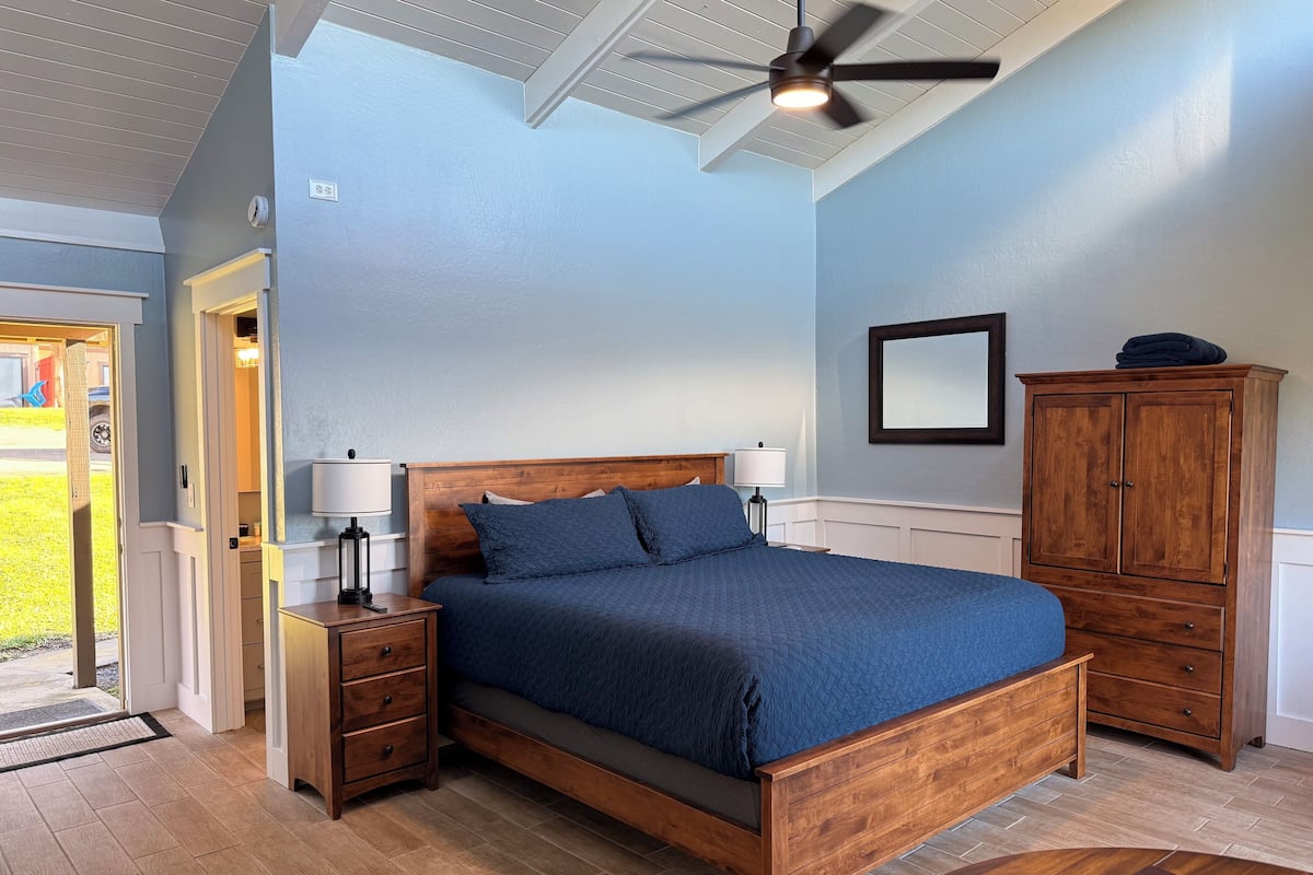 Quarto com cama de madeira, mesas de cabeceira, ventoinha no teto, guarda-roupa e porta que dá para uma varanda.