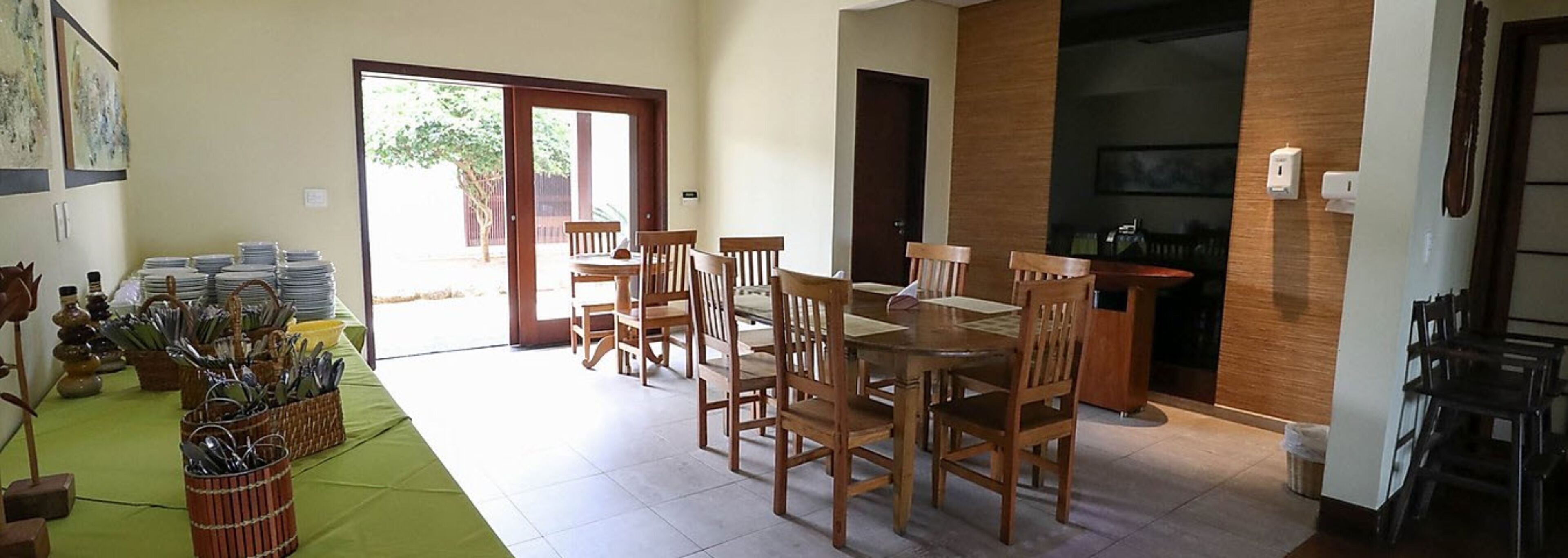 Uma sala de jantar com mesas e cadeiras de madeira, toalha de mesa verde e vista para o exterior através de uma porta de vidro.