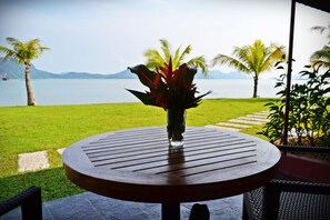 Uma mesa de madeira redonda com um vaso de flores vermelhas, colocada num pátio com vista para um lago calmo e montanhas ao longe.