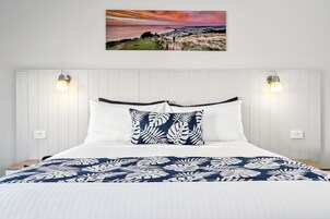 Uma cama com roupa de cama branca e azul, duas luminárias na parede e uma pintura de paisagem acima.