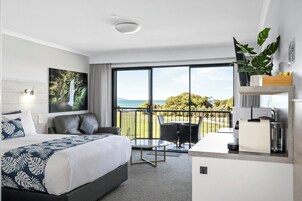 Quarto de hotel com cama, escrivaninha, cadeira, varanda e vista para o mar, além de um quadro na parede.