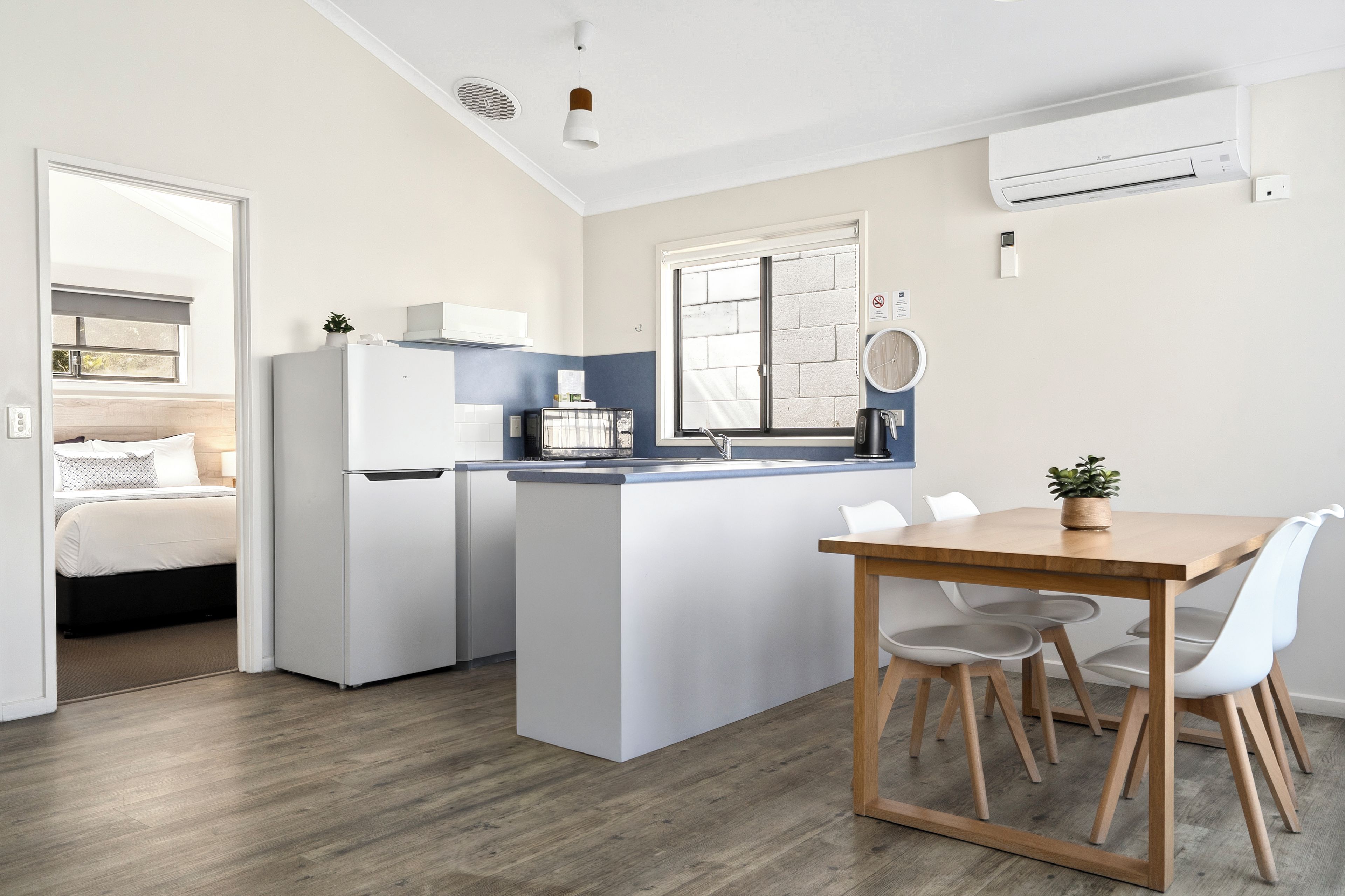 Cozinha compacta com ilha central, área de refeições com mesa e cadeiras de madeira, e uma janela visível.