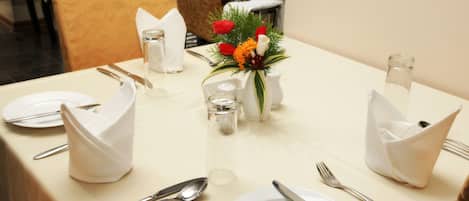 Mesa de jantar bem posta, com toalha branca, talheres e um arranjo floral.
