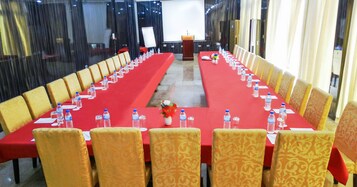 Uma sala de conferências com uma mesa comprida, cadeiras amarelas, garrafas de água e uma tela de projetor.