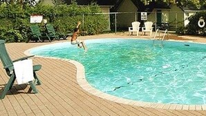 Uma piscina com deck revestido, rodeada por espreguiçadeiras e um pequeno edifício ao fundo.