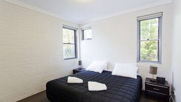 Quarto com edredom acolchoado preto, duas almofadas brancas, mesa de cabeceira com abajur e duas janelas com vista para o verde.