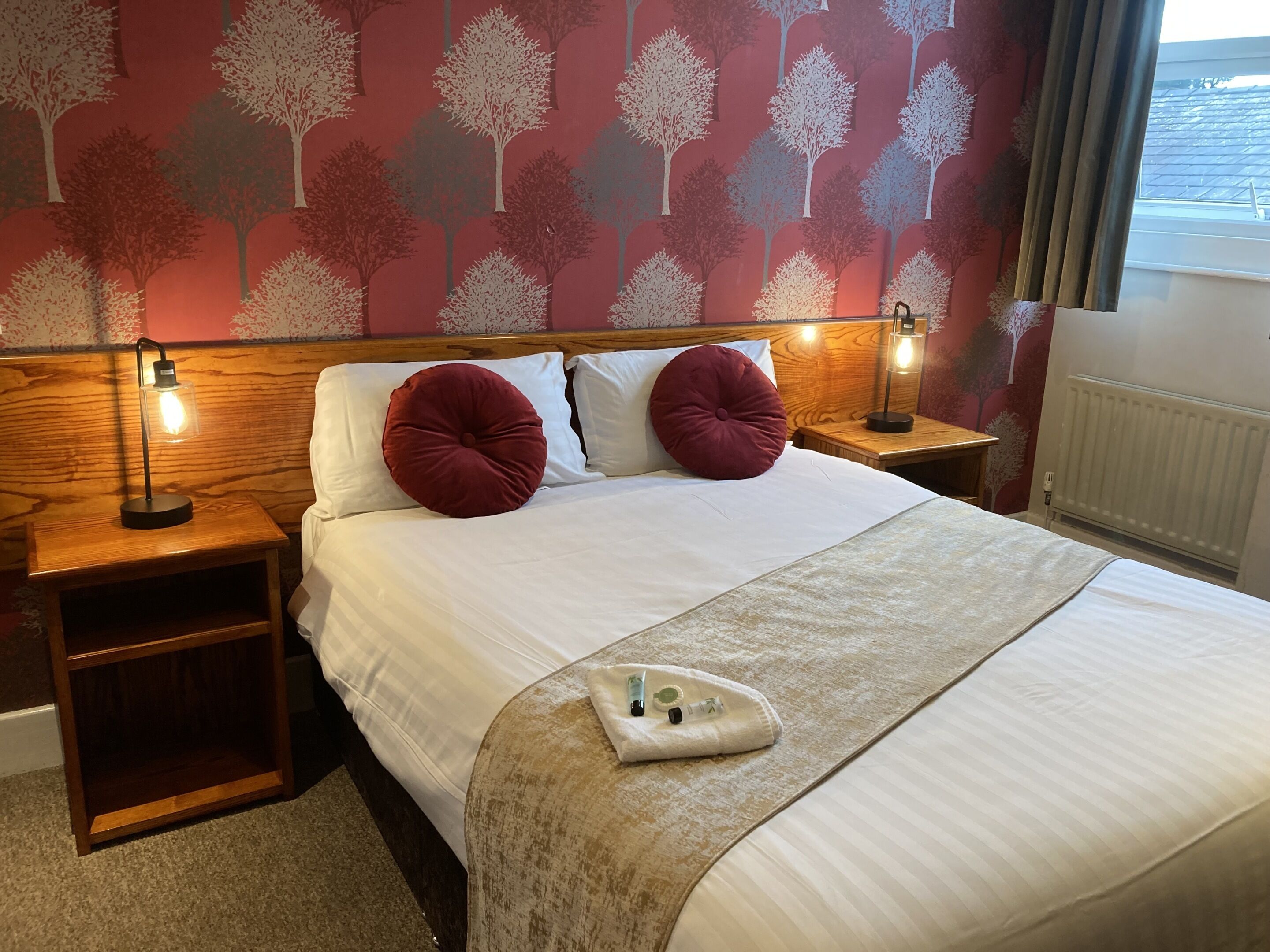 Quarto de hotel com cama, duas luminárias de cabeceira, papel de parede estampado em vermelho e branco e janela com cortinas.