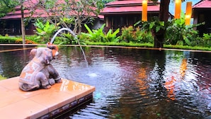Uma fonte com uma estátua de elefante, rodeada por vegetação exuberante e edifícios tradicionais com telhados vermelhos.