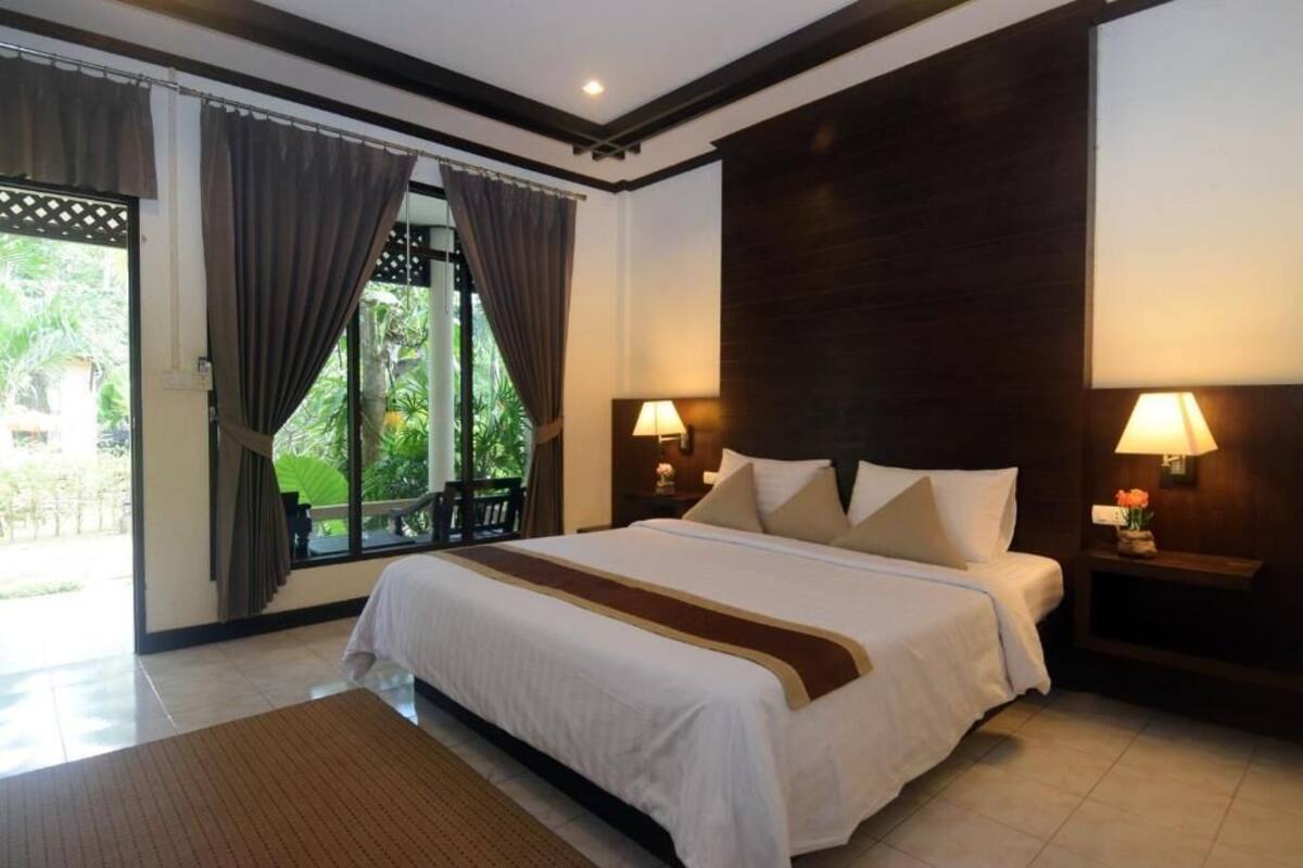 Quarto de hotel com uma cama grande, duas luminárias de cabeceira, vista para o verde pela janela e cabeceira de madeira.