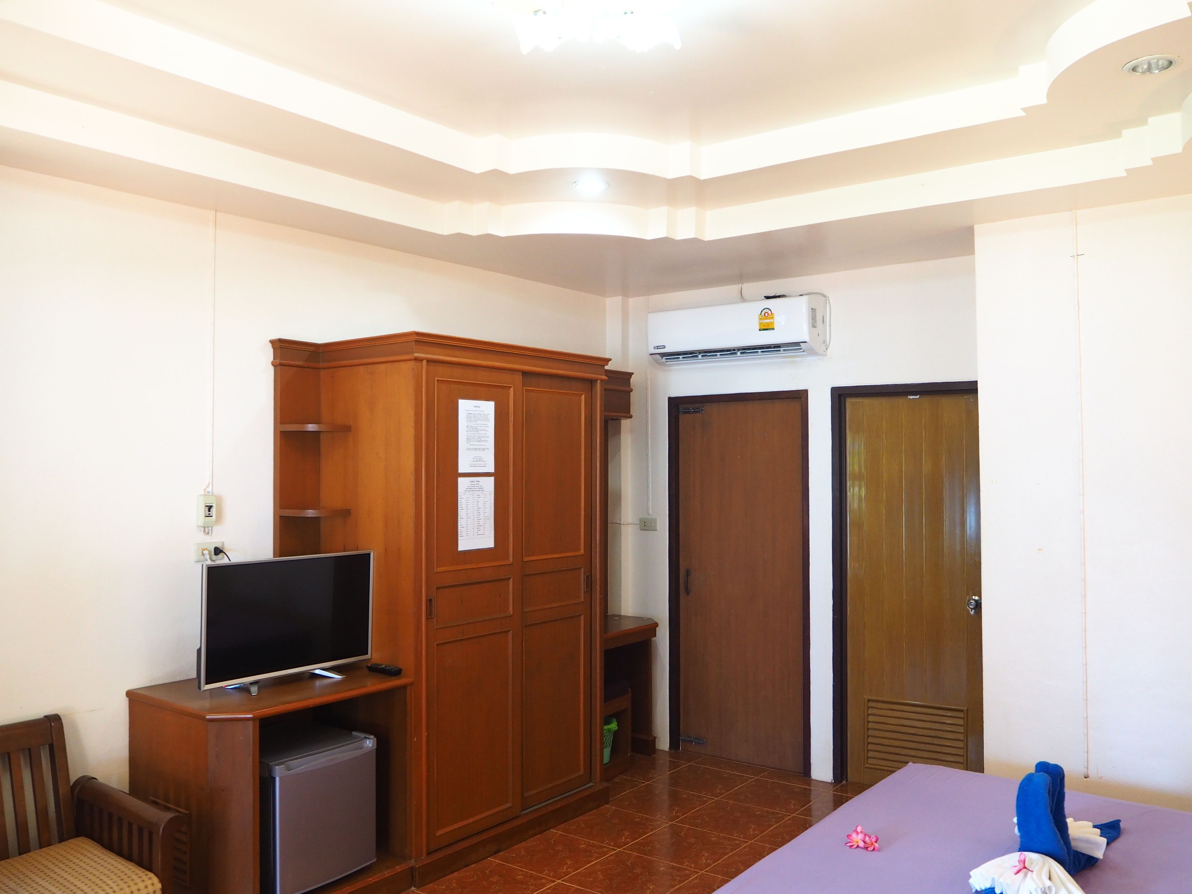 Quarto de hotel com armário de madeira, televisão, pequeno frigorífico, cama com colcha roxa e unidade de ar condicionado na parede.