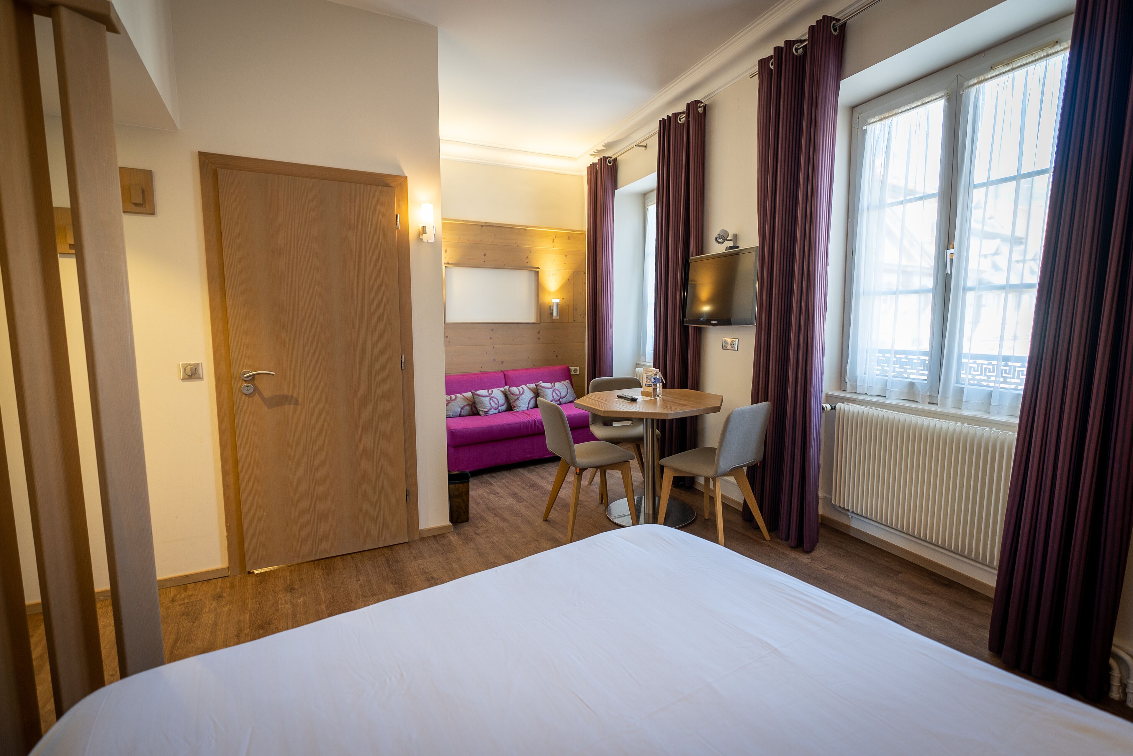 Quarto de hotel com cama, sofá, mesa de jantar com cadeiras, televisão e janela com cortinas.
