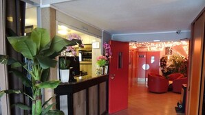 Um hall de hotel com balcão de recepção, plantas em vasos, uma porta vermelha e uma área de estar com cadeiras vermelhas.