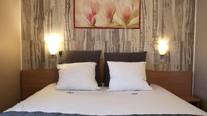Quarto de hotel com cama, duas almofadas brancas, edredom acolchoado escuro e quadro com flores rosa na parede.