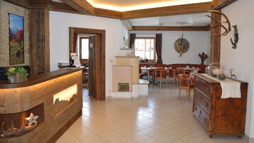 Um saguão de hotel com um balcão de recepção de madeira, uma lareira, uma área de refeições com mesas e cadeiras, e uma grande janela com cortinas.