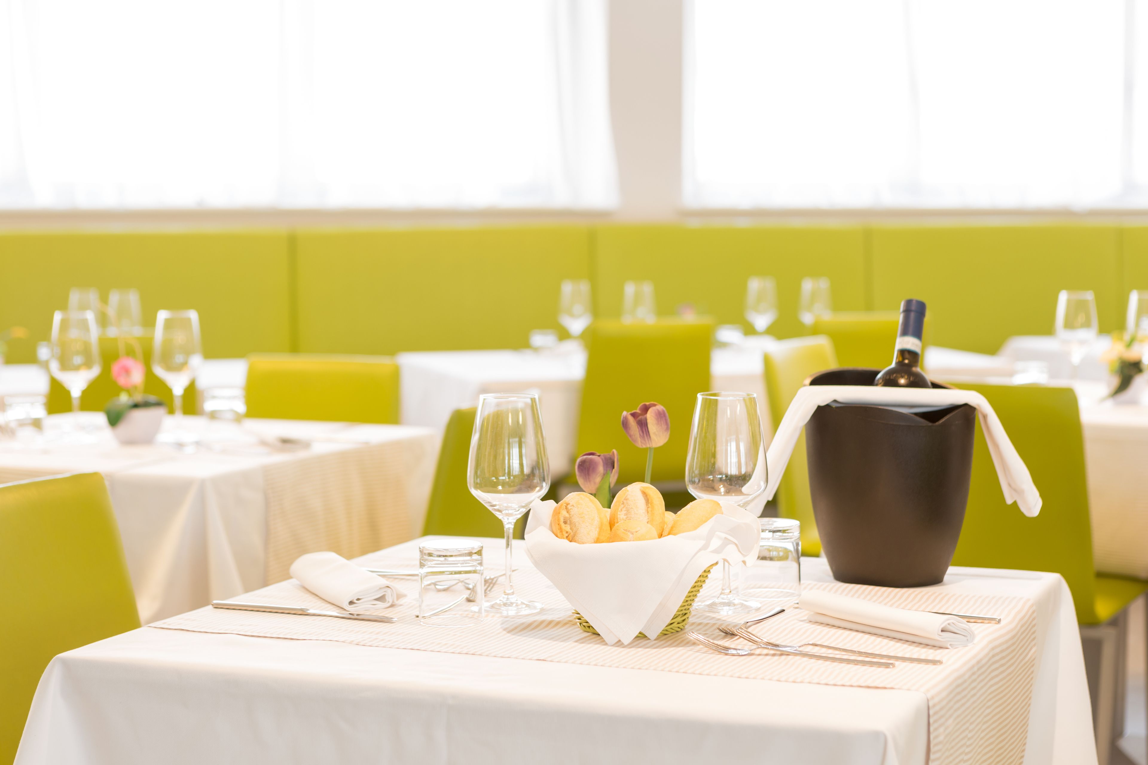 Ambiente de restaurante com mesas preparadas para refeições, incluindo copos de vinho, cestas de pão e uma garrafa de vinho.
