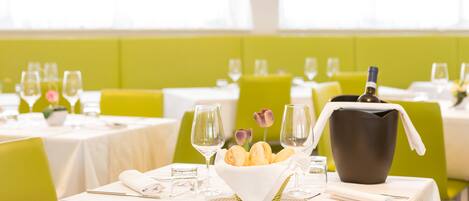 Ambiente de restaurante com mesas preparadas para refeições, incluindo copos de vinho, cestas de pão e uma garrafa de vinho.