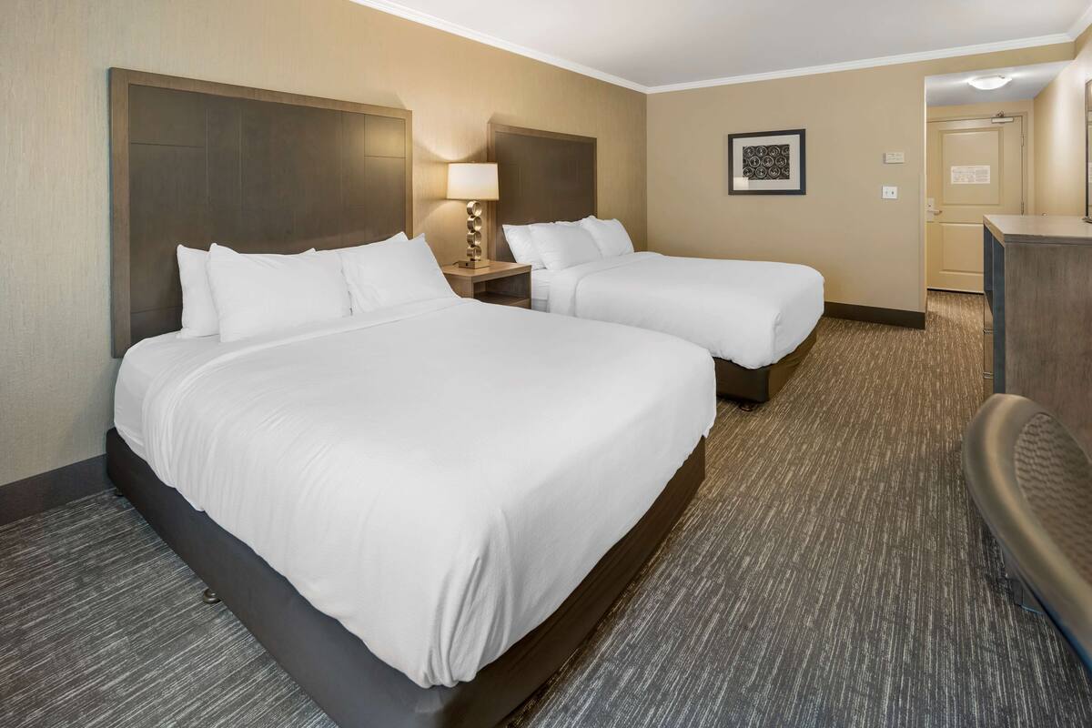 Quarto de hotel com duas camas, uma escrivaninha, um abajur e uma porta.