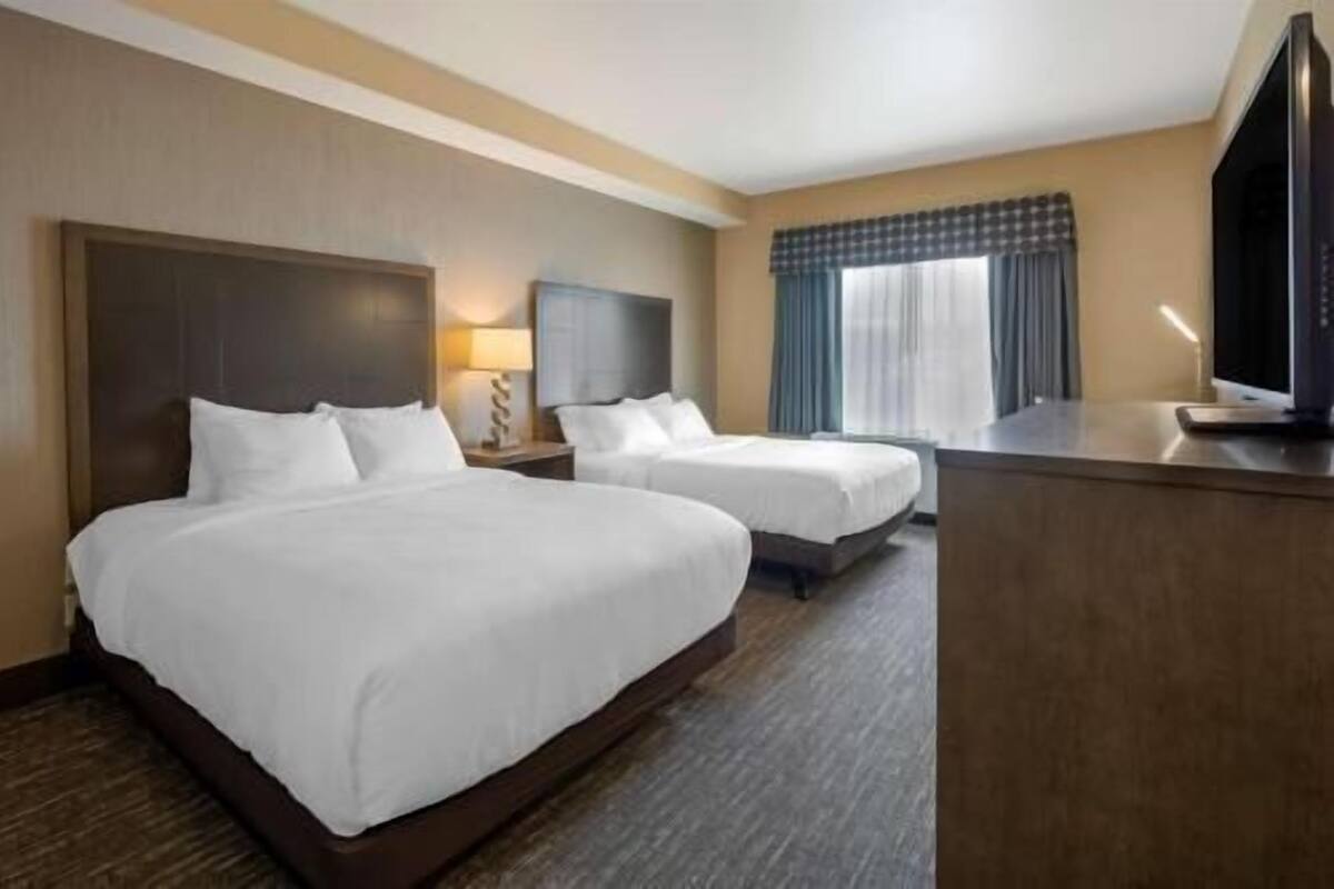 Quarto de hotel com duas camas, uma escrivaninha, televisão e janela com cortinas.