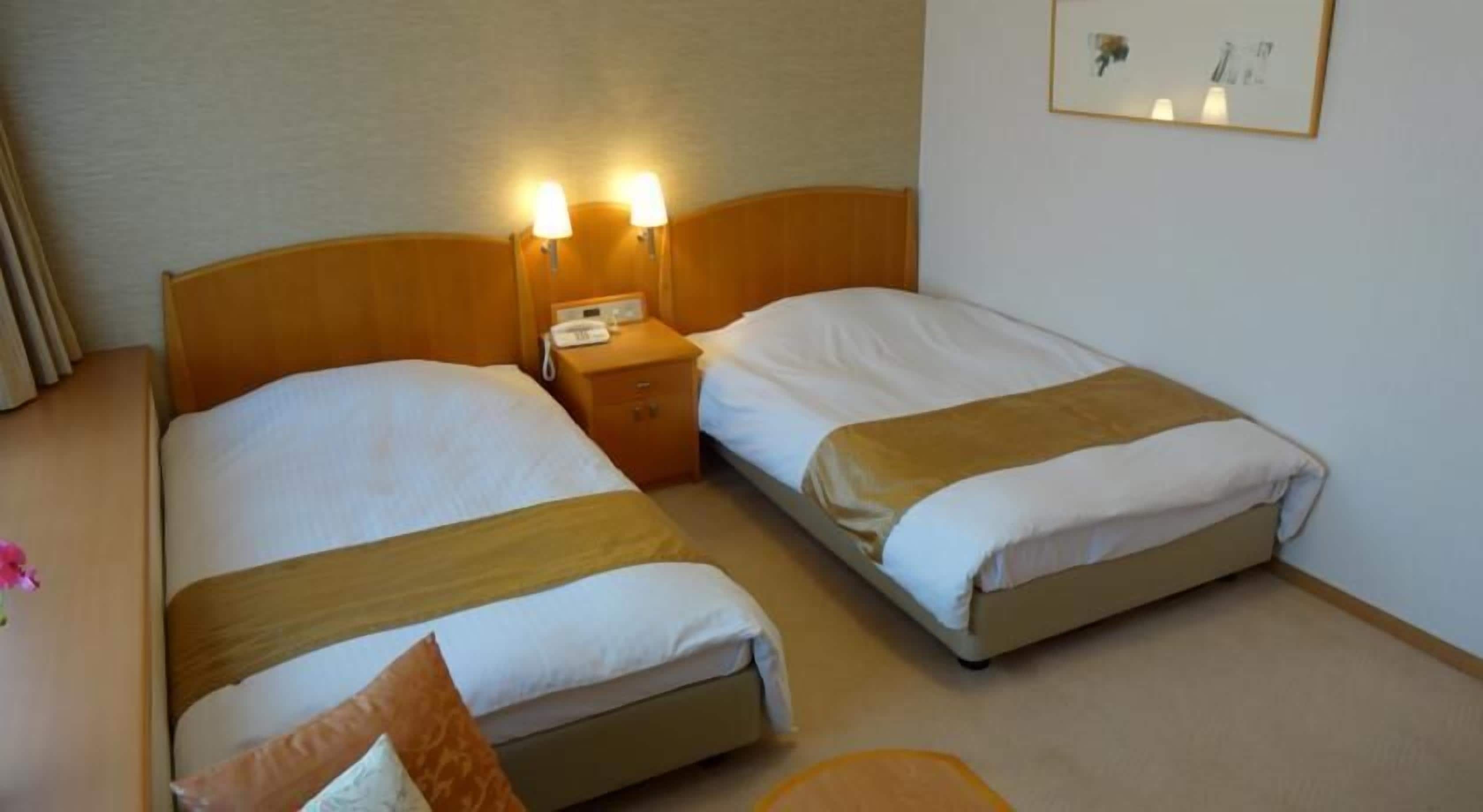 Quarto de hotel com duas camas de solteiro, uma mesa de cabeceira com abajur e uma pequena mesa com telefone.