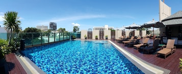 Uma piscina no terraço com água azul cristalina, cercada por espreguiçadeiras e guarda-sóis, com edifícios da cidade ao fundo.
