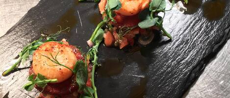 Uma tábua de ardósia preta com três vieiras, cada uma coberta com uma fatia de tomate e decorada com microverdes.