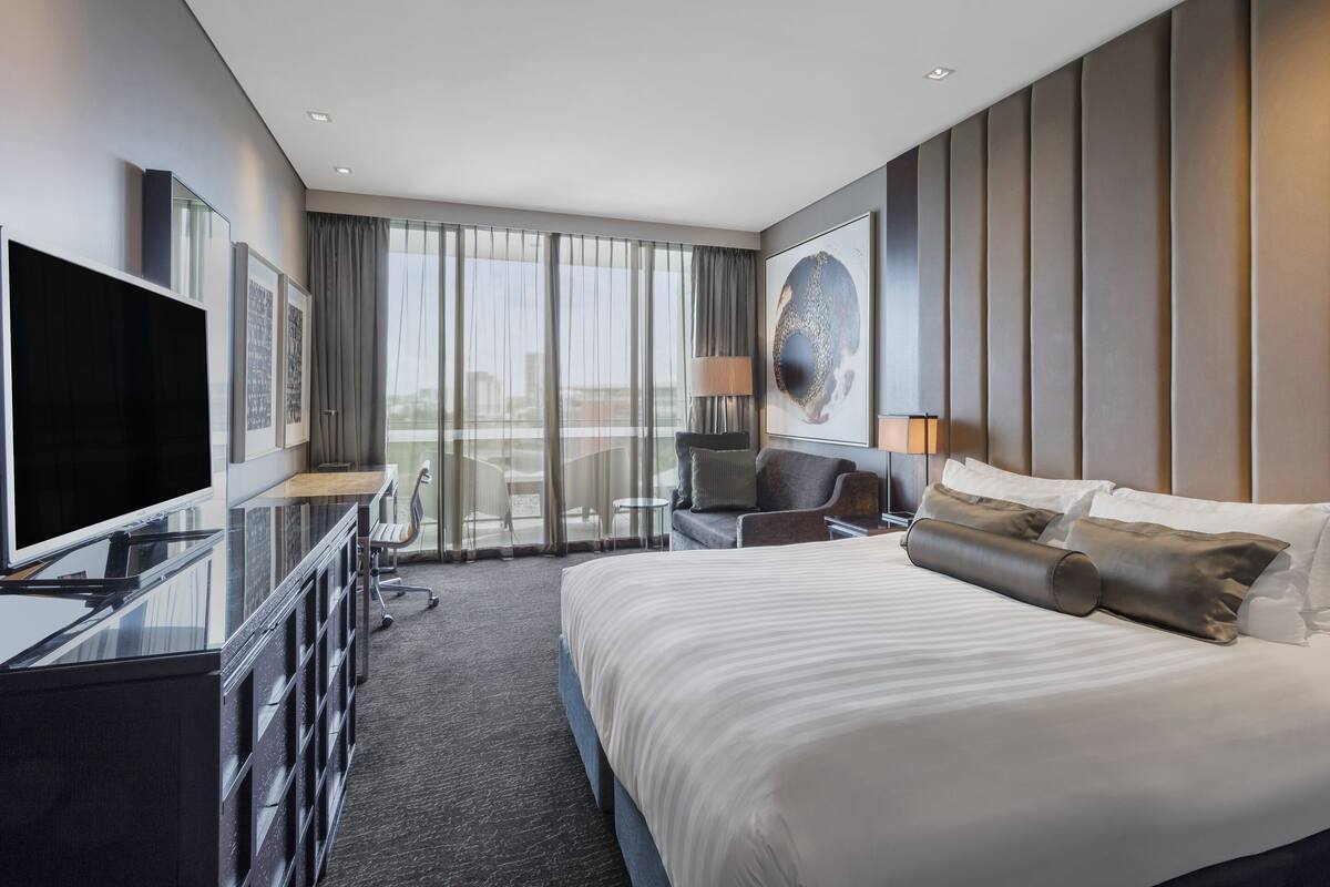 Quarto de hotel moderno com uma cama grande, uma escrivaninha com televisão de tela plana, um sofá e uma janela ampla com vista para a cidade.