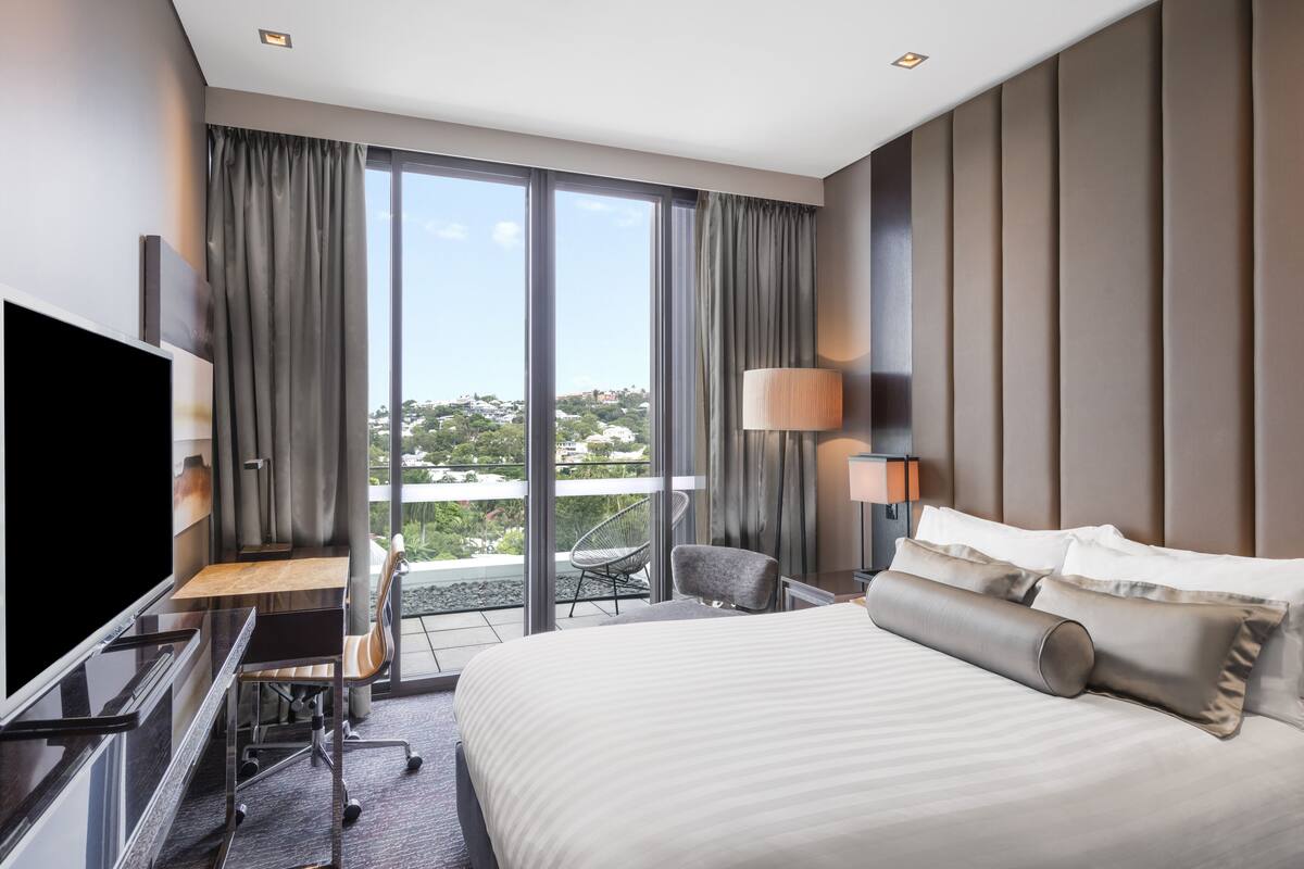 Quarto de hotel moderno com uma cama grande, uma escrivaninha com cadeira, televisão e varanda com vista para a cidade.
