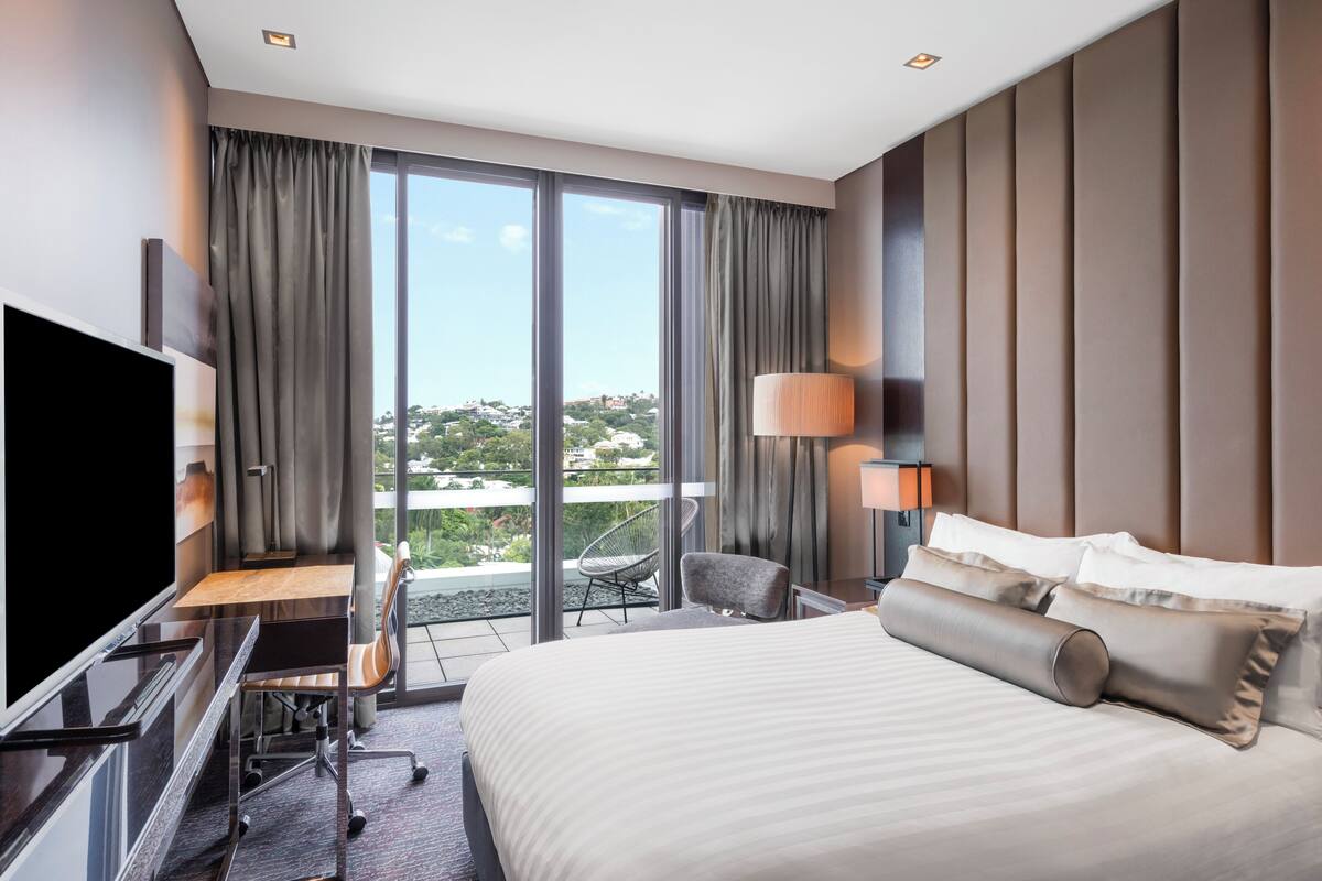 Quarto de hotel com uma cama grande, uma escrivaninha com cadeira, televisão e vista para a cidade através de amplas janelas.
