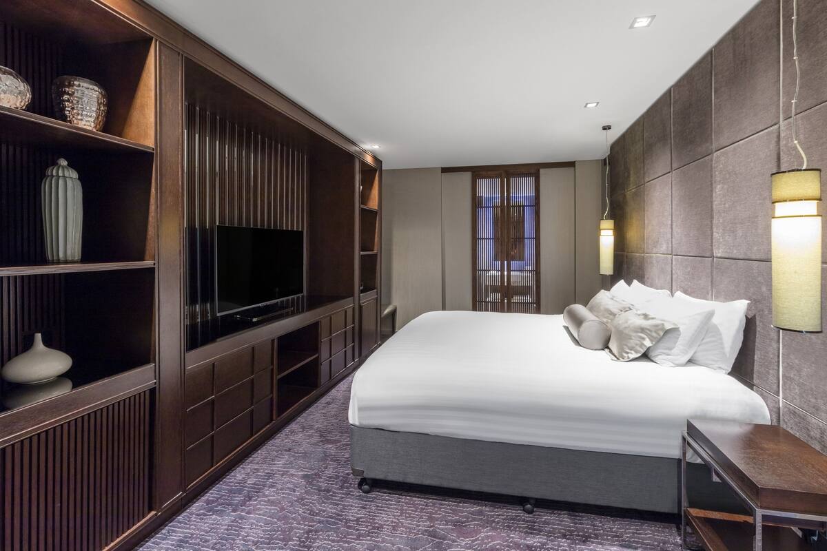 Quarto de hotel moderno com uma cama grande, TV integrada, estantes de madeira e uma janela ampla.