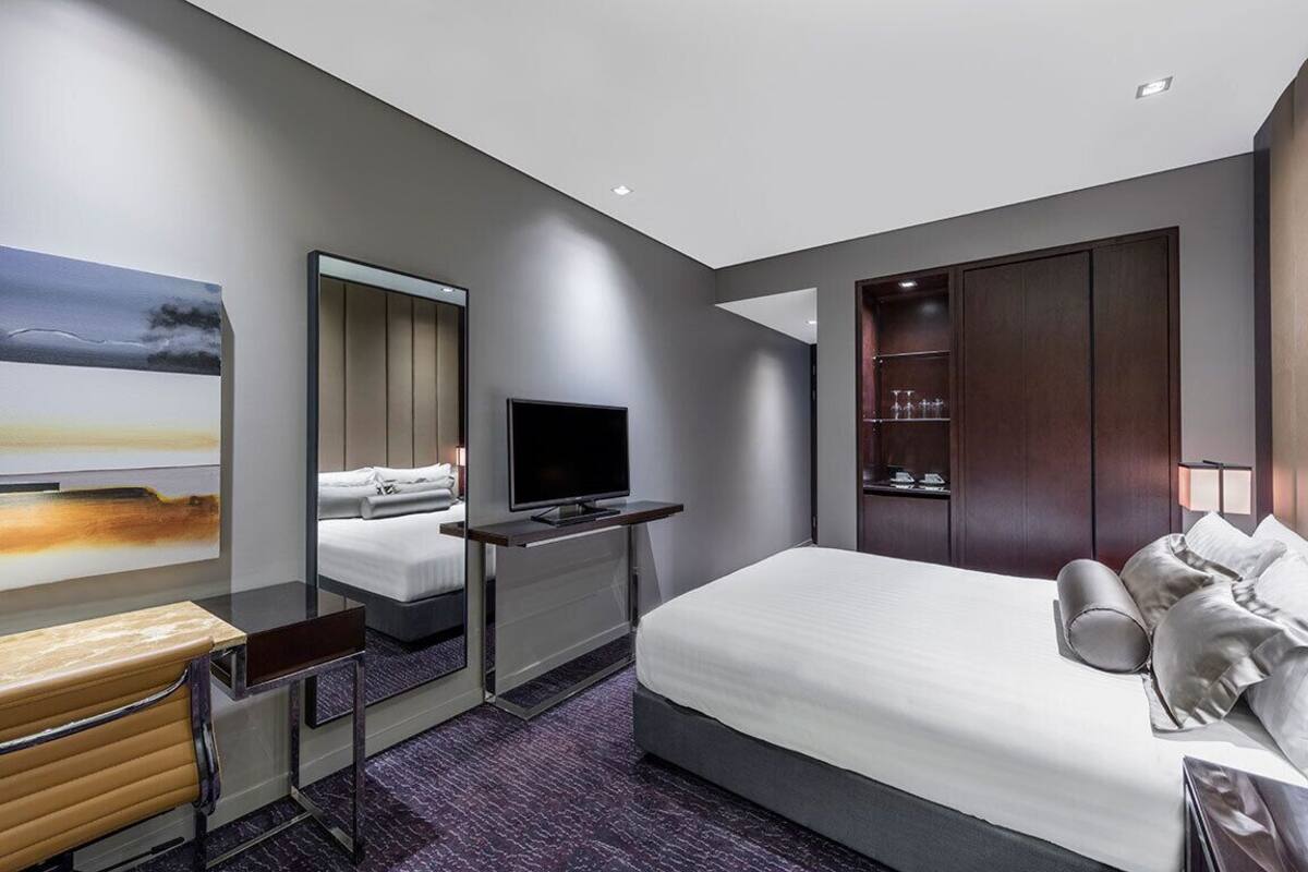 Quarto de hotel moderno com uma cama grande, televisão de tela plana, escrivaninha com cadeira, guarda-roupa e um quadro na parede.