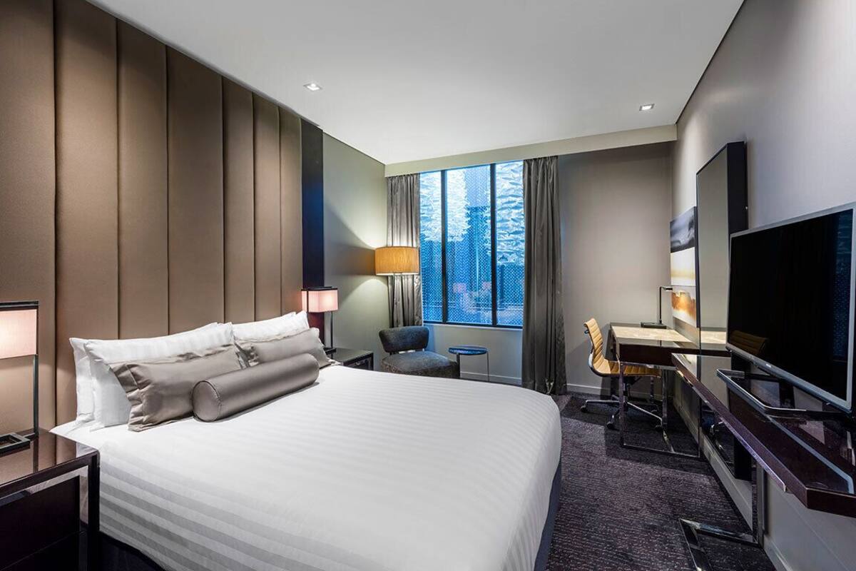 Quarto de hotel moderno com uma cama grande, uma escrivaninha, uma cadeira, televisão e janela com vista para árvores.