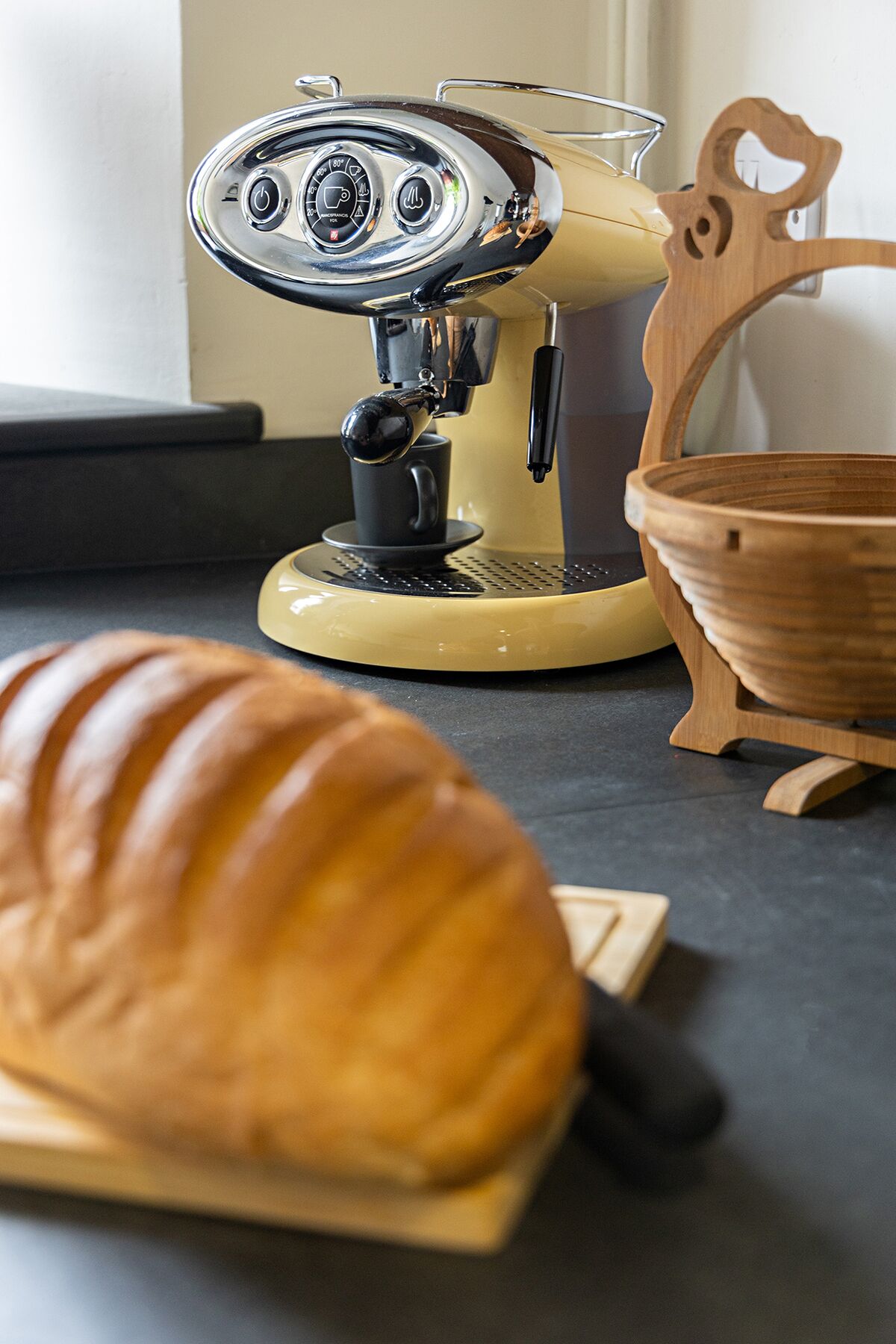 Uma máquina de café, uma cesta de pão de madeira e uma barra de pão em uma bancada.