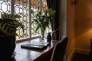 Uma mesa de madeira com um laptop, uma planta em vaso e um vaso com flores perto de uma janela com padrão geométrico.