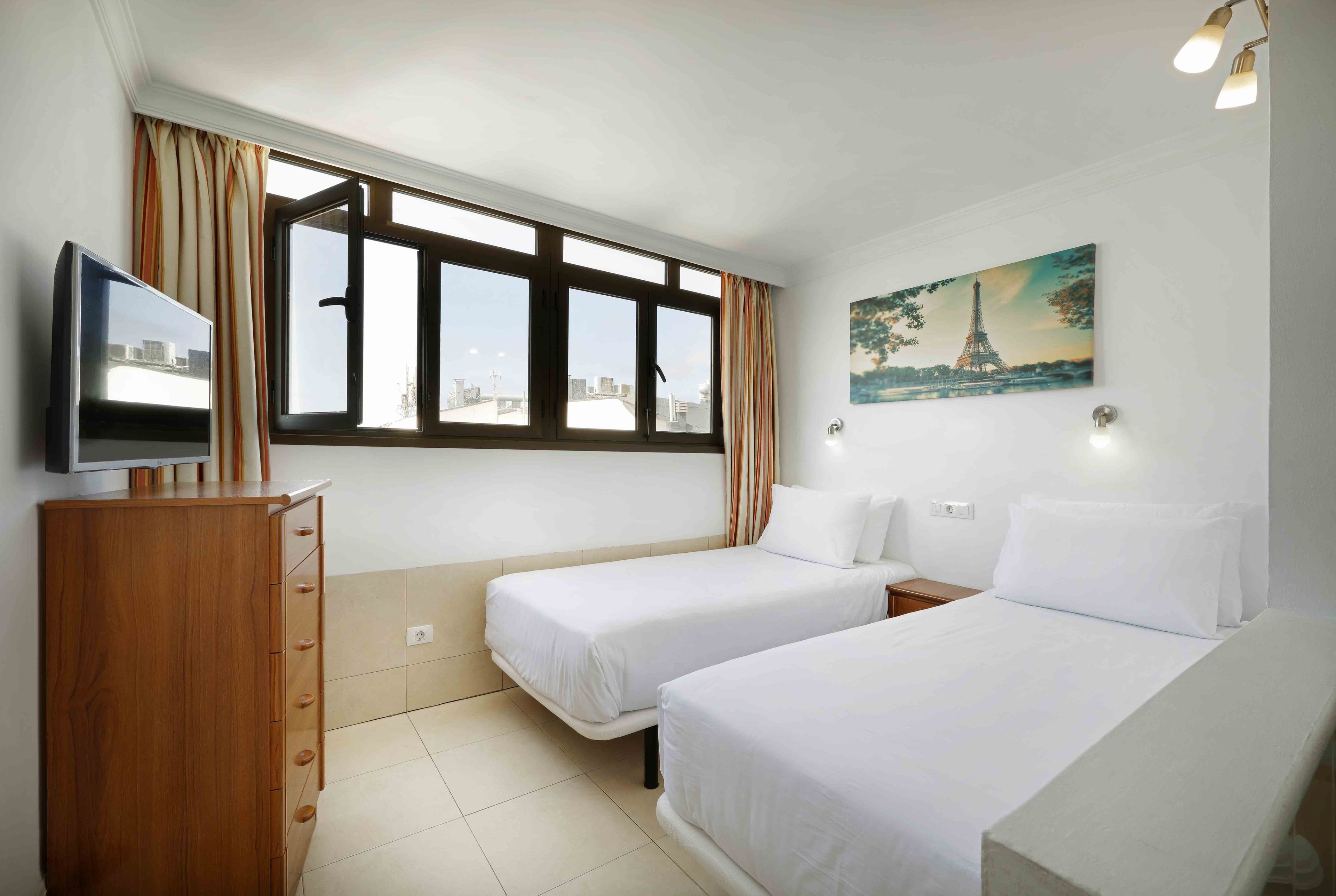 Quarto de hotel com duas camas de solteiro, cómoda de madeira, televisão de tela plana, janela com cortinas e quadro da Torre Eiffel.