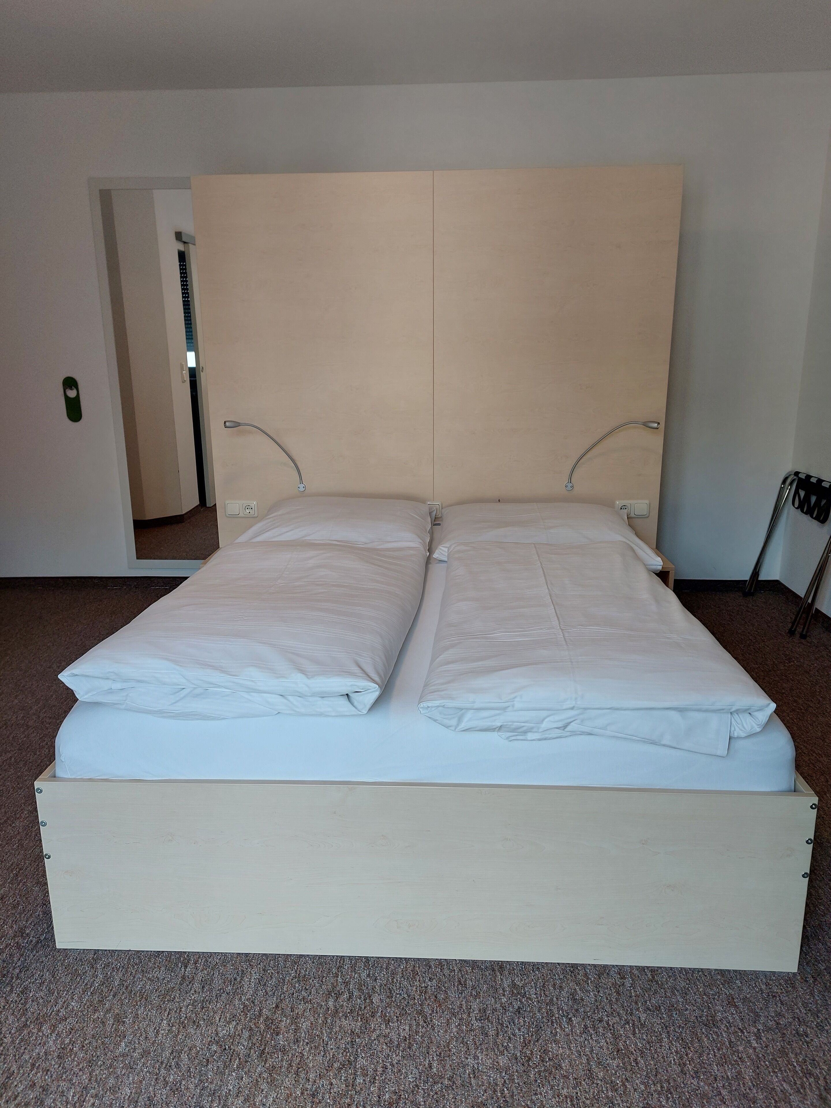 Uma cama de casal com estrutura e cabeceira de madeira, piso coberto por carpete e porta que dá acesso a outro quarto.