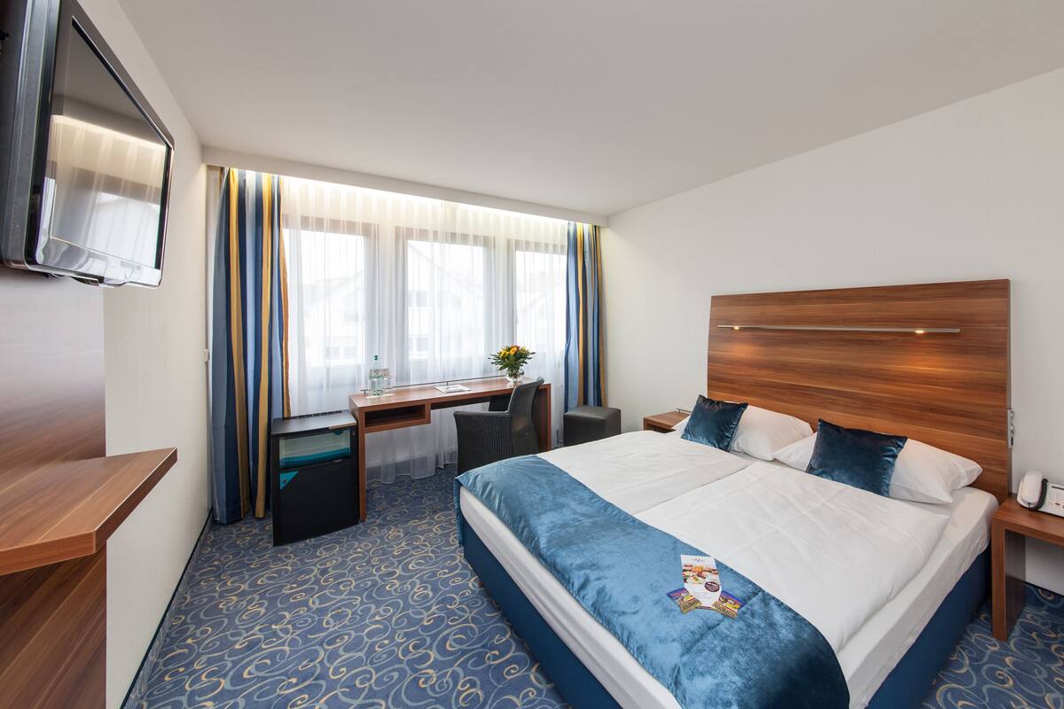 Quarto de hotel com uma cama grande, uma escrivaninha com cadeira, televisão na parede e uma janela com cortinas.