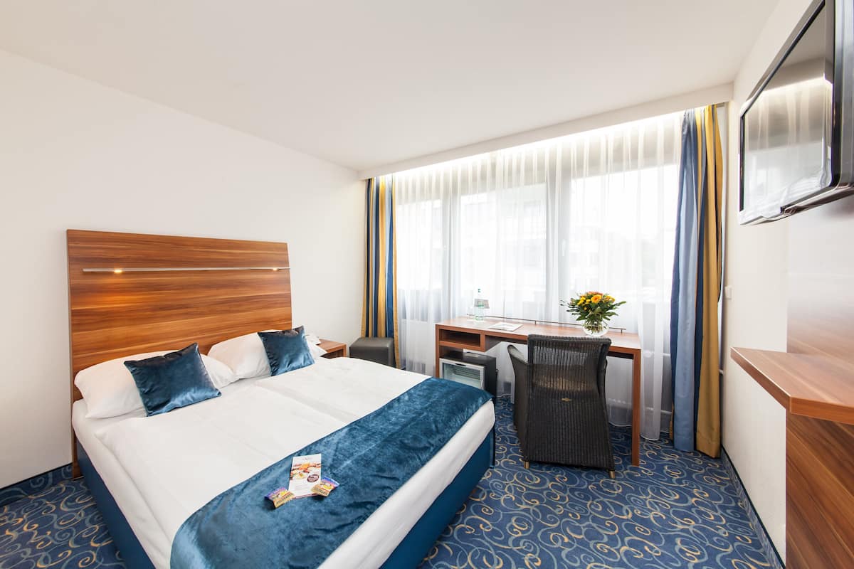 Quarto de hotel com uma cama grande, uma escrivaninha, uma cadeira, uma televisão e uma janela com cortinas.