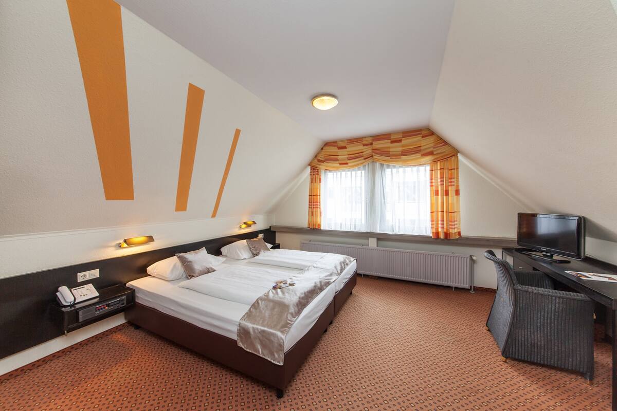 Quarto de hotel com cama, escrivaninha, televisão e janela com cortinas.