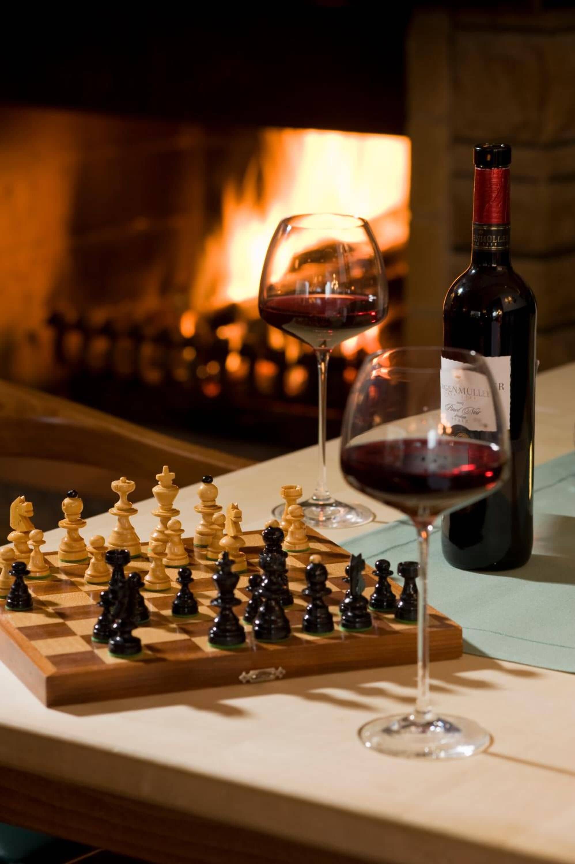 Um tabuleiro de xadrez com peças, dois copos de vinho e uma garrafa de vinho sobre uma mesa, com uma lareira ao fundo.