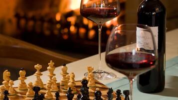 Um tabuleiro de xadrez com peças, dois copos de vinho e uma garrafa de vinho sobre uma mesa, com uma lareira ao fundo.