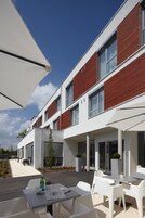 Um edifício moderno com telhado plano, janelas amplas e uma área externa coberta com mesas e cadeiras brancas.