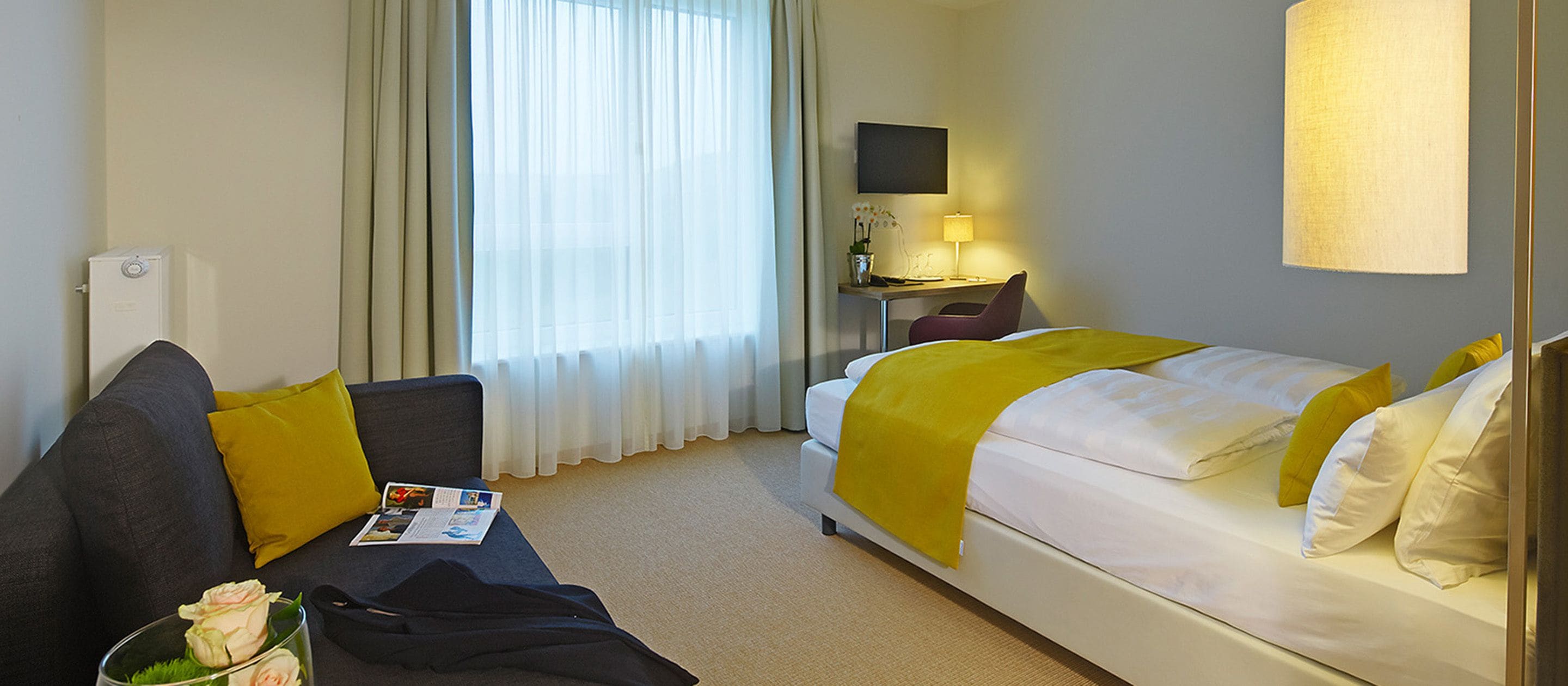 Quarto de hotel com cama, sofá, escrivaninha com luminária e televisão.